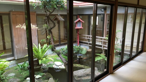 Um quarto em estilo japonês tradicional com tatames, portas deslizantes e vista para um jardim com lago, pedras e uma estrutura de madeira.