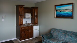 Cozinha compacta com armários de madeira, micro-ondas e um refrigerador pequeno. Na sala de estar, há um sof á estampado e um quadro na parede.