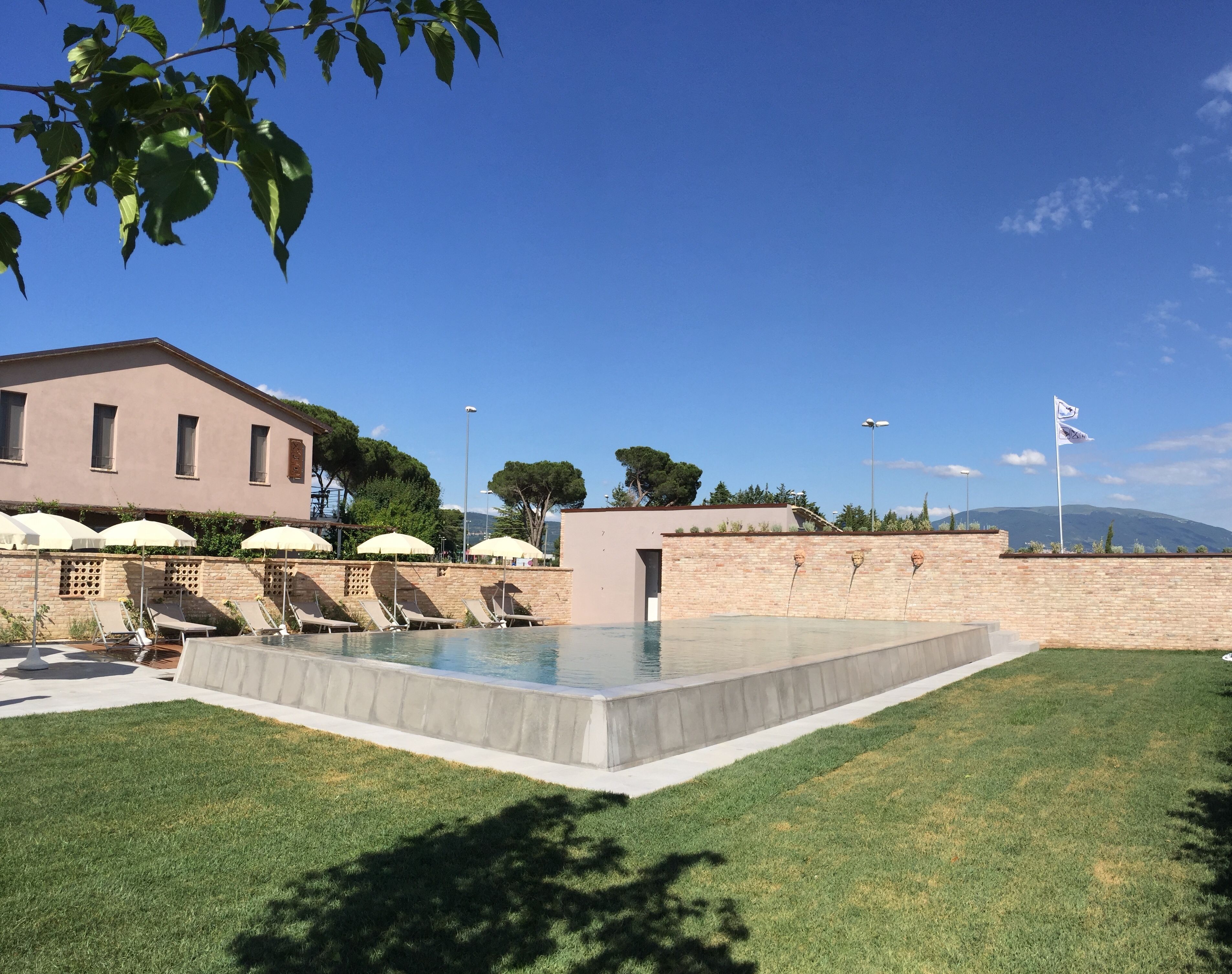 Uma área de piscina com espreguiçadeiras e guarda-sóis, uma parede de tijolos e um edifício ao fundo.