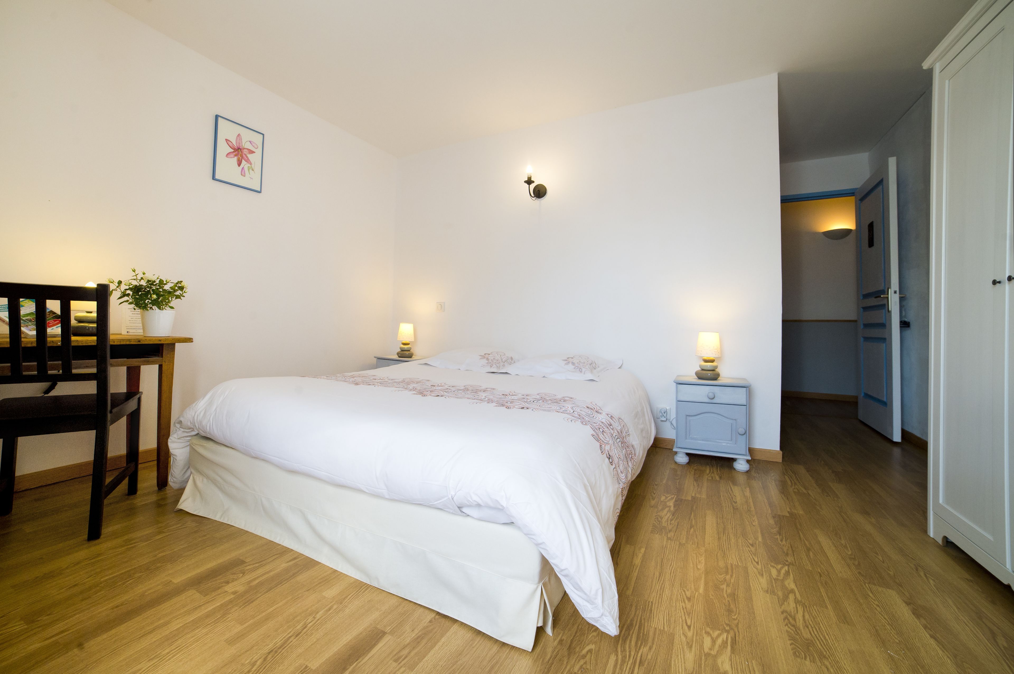 Quarto com cama, mesas de cabeceira, uma cadeira e porta que dá para outro cômodo.