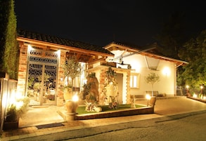 Edifício residencial bem iluminado com telhado em telhas, entrada com portão e um jardim frontal ajardinado com elementos decorativos.