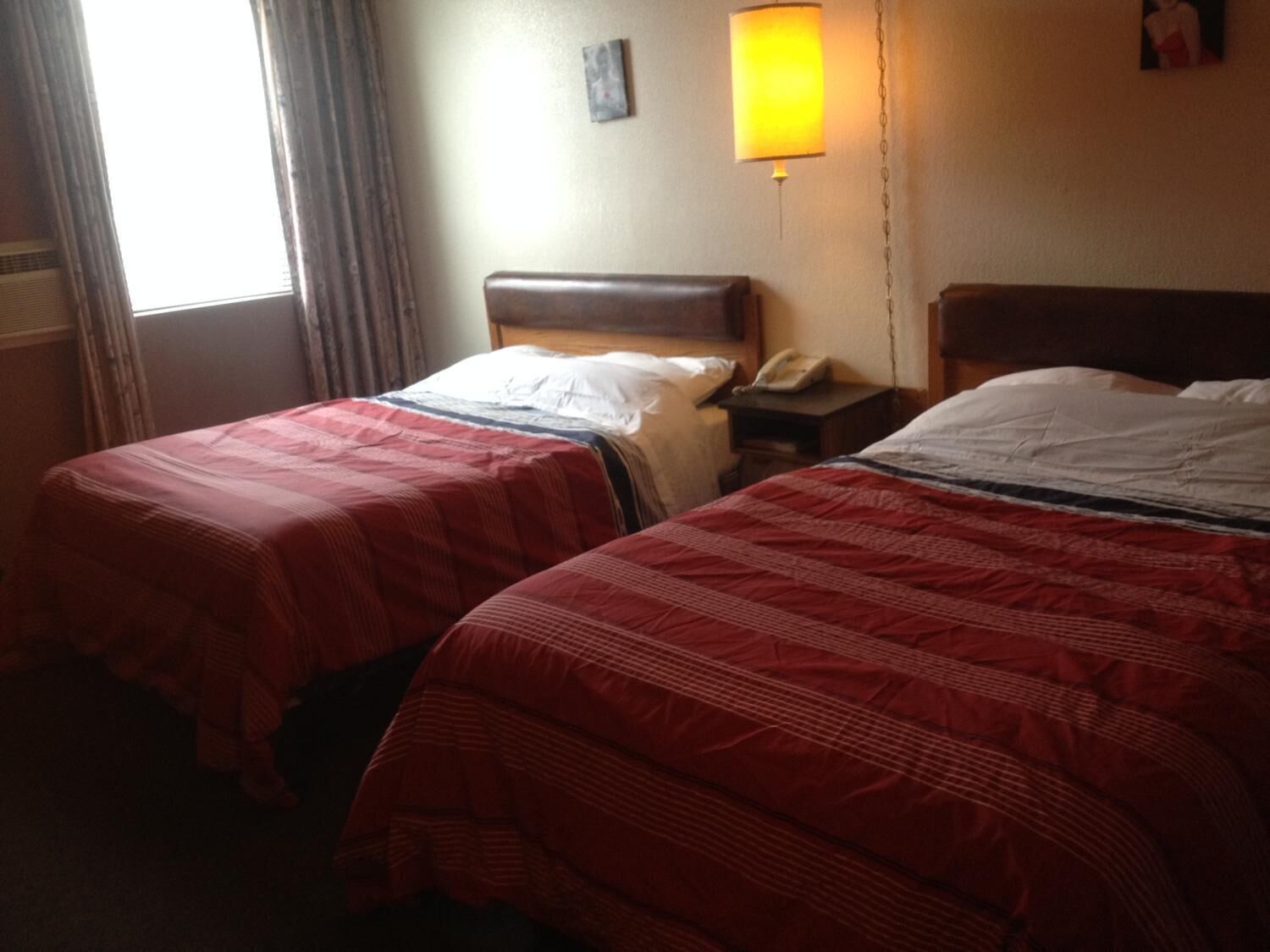 Quarto de hotel com duas camas de solteiro, uma mesinha de cabeceira com abajur e uma janela com cortinas.