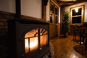 Uma lareira a lenha com porta de vidro, parede de tijolos, soalho de madeira, um vaso com planta e uma janela com luminária suspensa.
