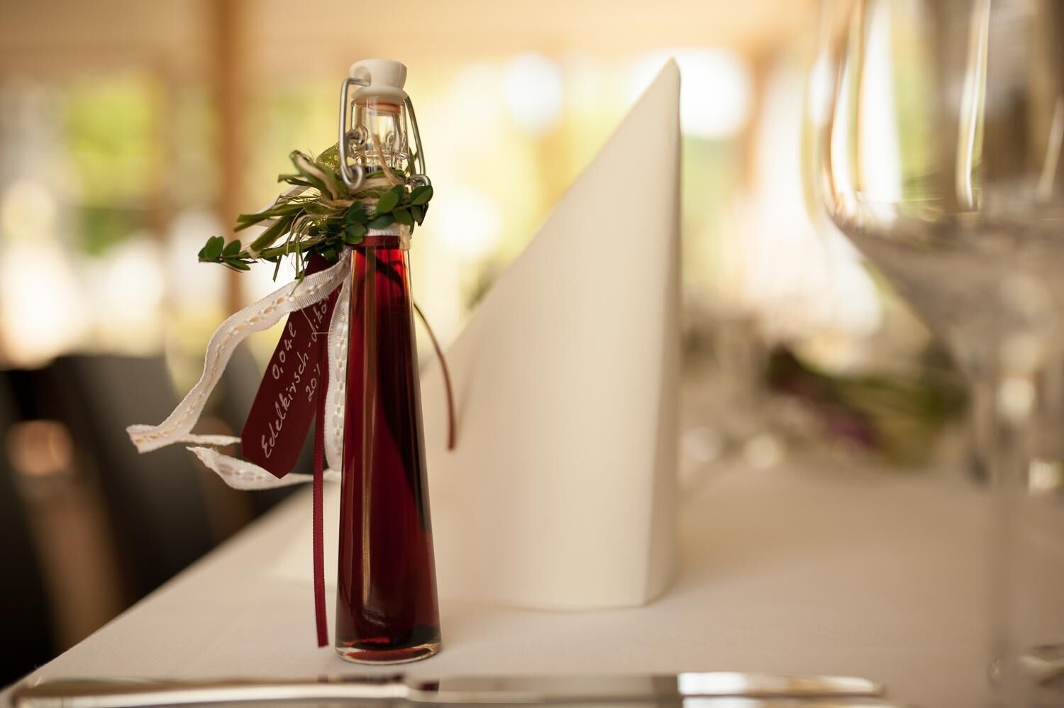 Uma garrafa de líquido vermelho com etiqueta branca e um laço, colocada sobre uma mesa com um guardanapo branco e um copo.