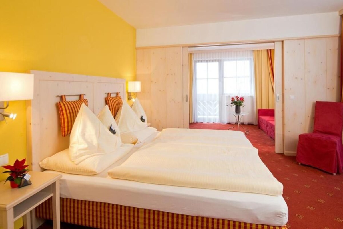 Quarto de hotel com cama grande, mesa de cabeceira, uma cadeira vermelha e janela com cortinas.