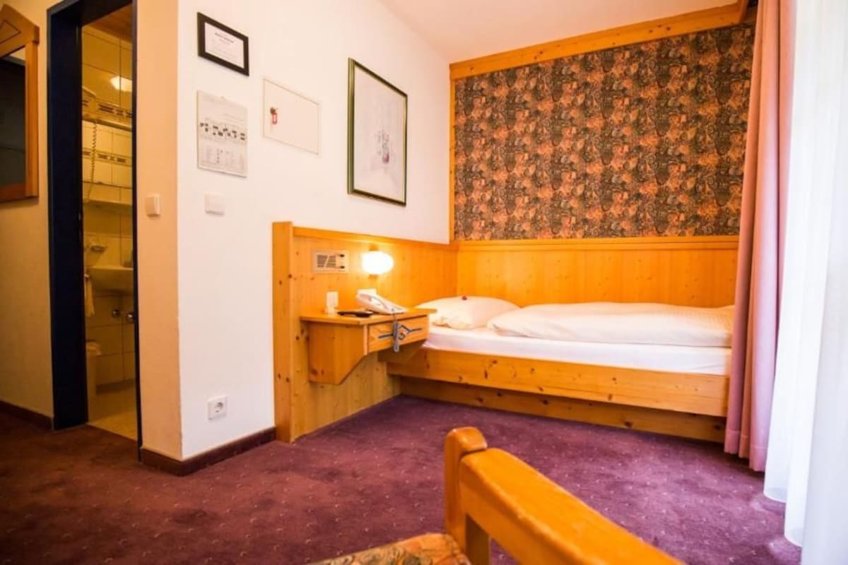 Quarto de hotel com uma cama de solteiro, cabeceira de madeira, mesa de cabeceira com abajur e telefone fixado na parede.