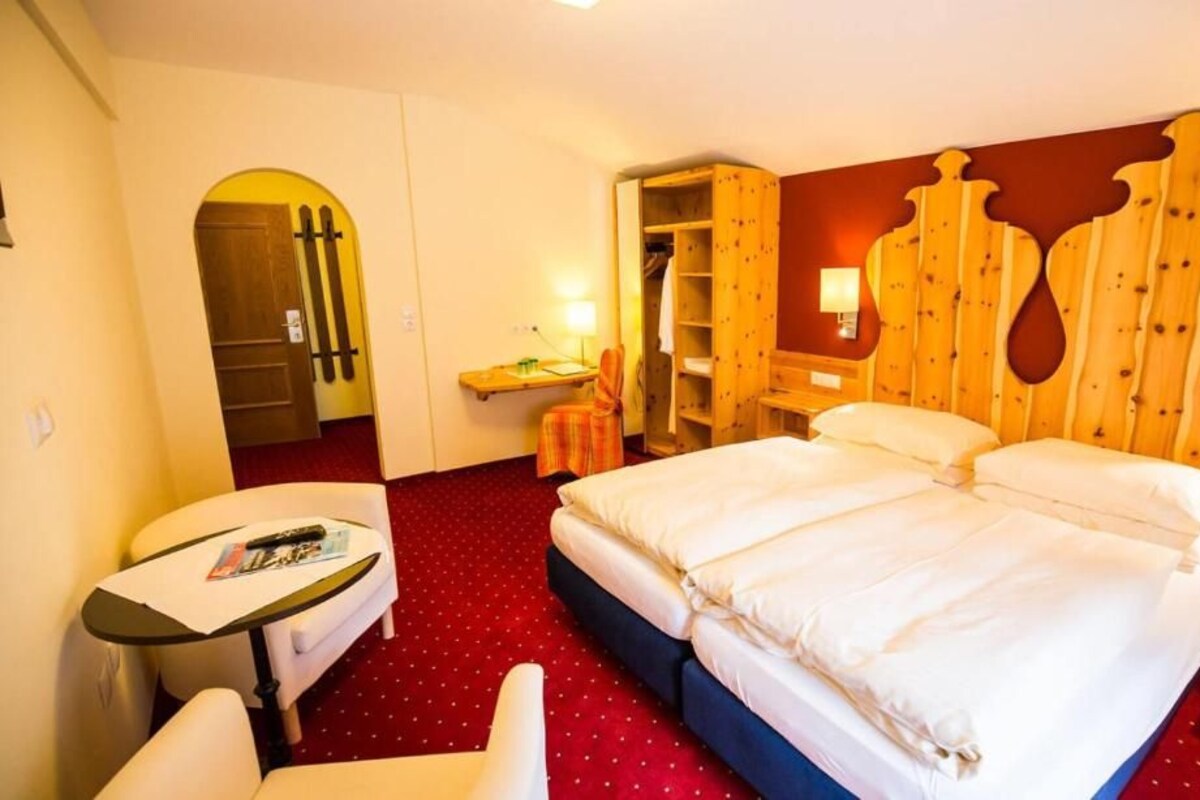 Quarto de hotel com cama de casal, uma pequena mesa com uma revista, uma cadeira, um guarda-roupa e um espelho.
