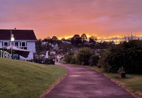 Rua residencial ao entardecer, com casas, céu limpo e gramado bem cuidado.