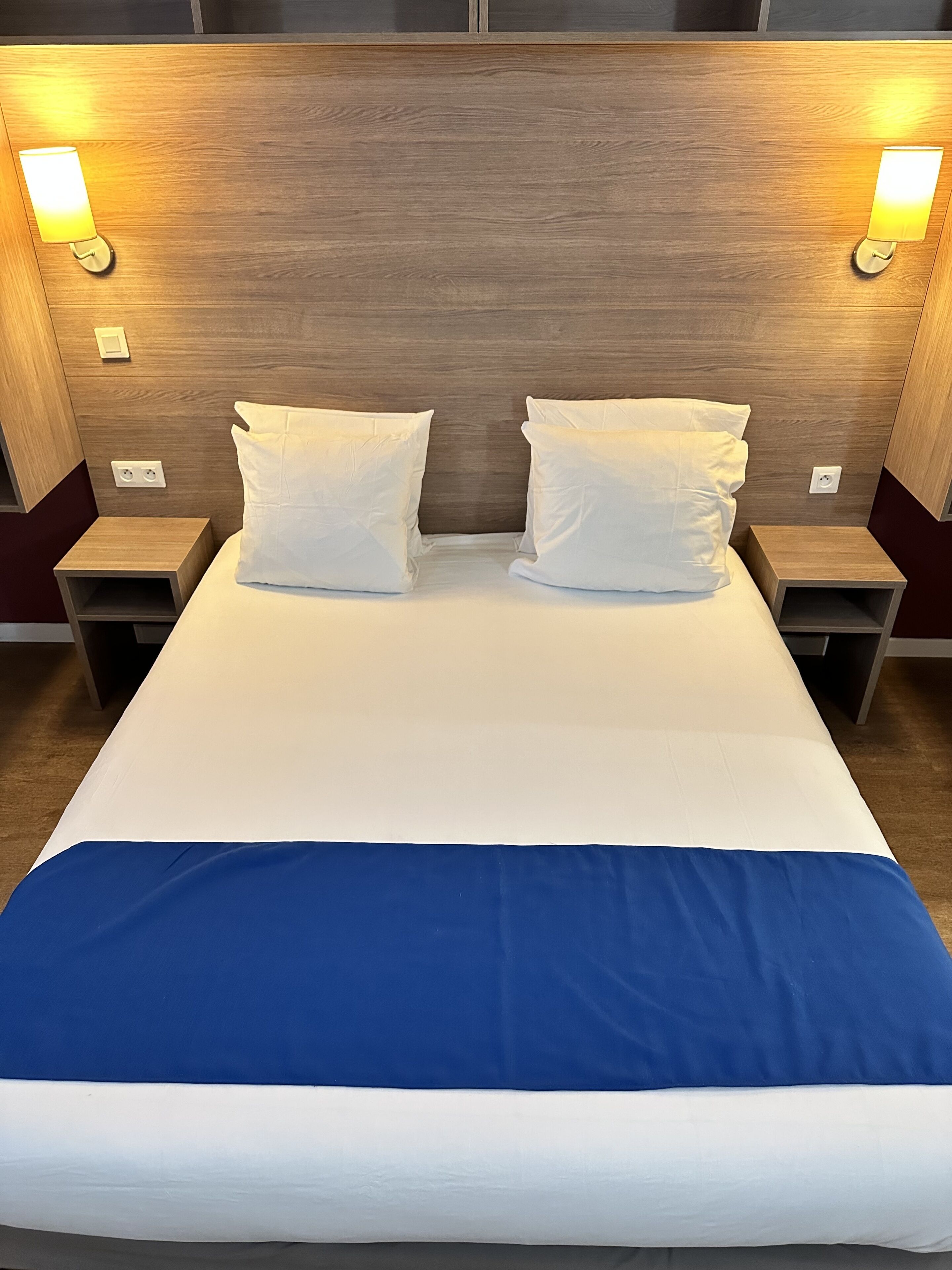 Quarto de hotel com cabeceira de madeira, duas mesas de cabeceira com luminárias, cama com roupa de cama branca e azul, e almofadas brancas.