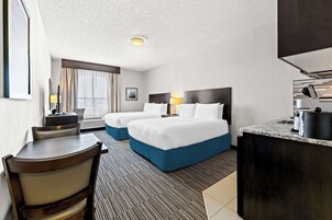 Quarto de hotel com duas camas, uma escrivaninha, uma cadeira, uma mesinha, uma luminária e uma cozinha compacta.