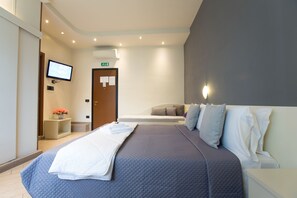Quarto de hotel moderno com uma cama grande, uma TV de tela plana na parede, uma pequena mesa de cabeceira com um vaso de flores e um sofá-cama encostado na parede.