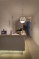 Interior moderno com uma ilha central que possui uma televisão, um candeeiro branco e uma planta em vaso. Uma escadaria é visível ao fundo.
