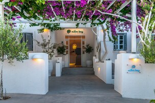 Entrada de hotel com fachada branca, placa indicando "Milos Bay Suites" e galeria coberta com flores roxas.