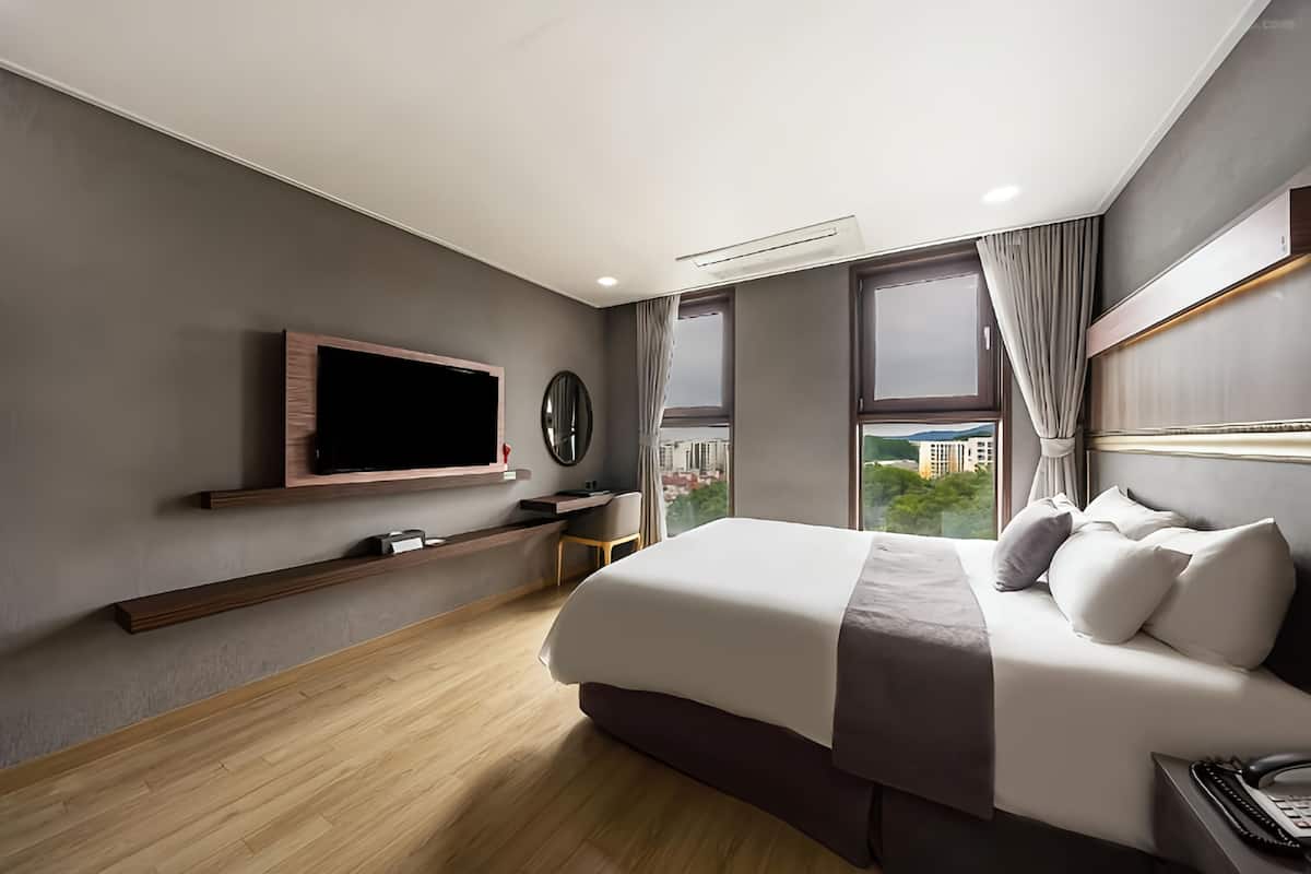 Quarto de hotel moderno com uma cama grande, uma televisão de tela plana na parede, uma escrivaninha com uma cadeira e vista para a cidade através de grandes janelas.