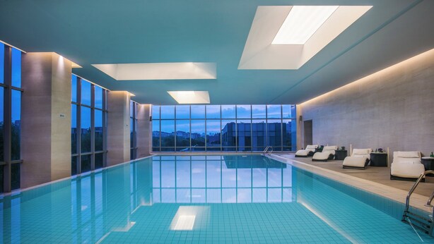 Piscina interior moderna com água azul cristalina, amplas janelas e painéis de teto iluminados pelo sol.