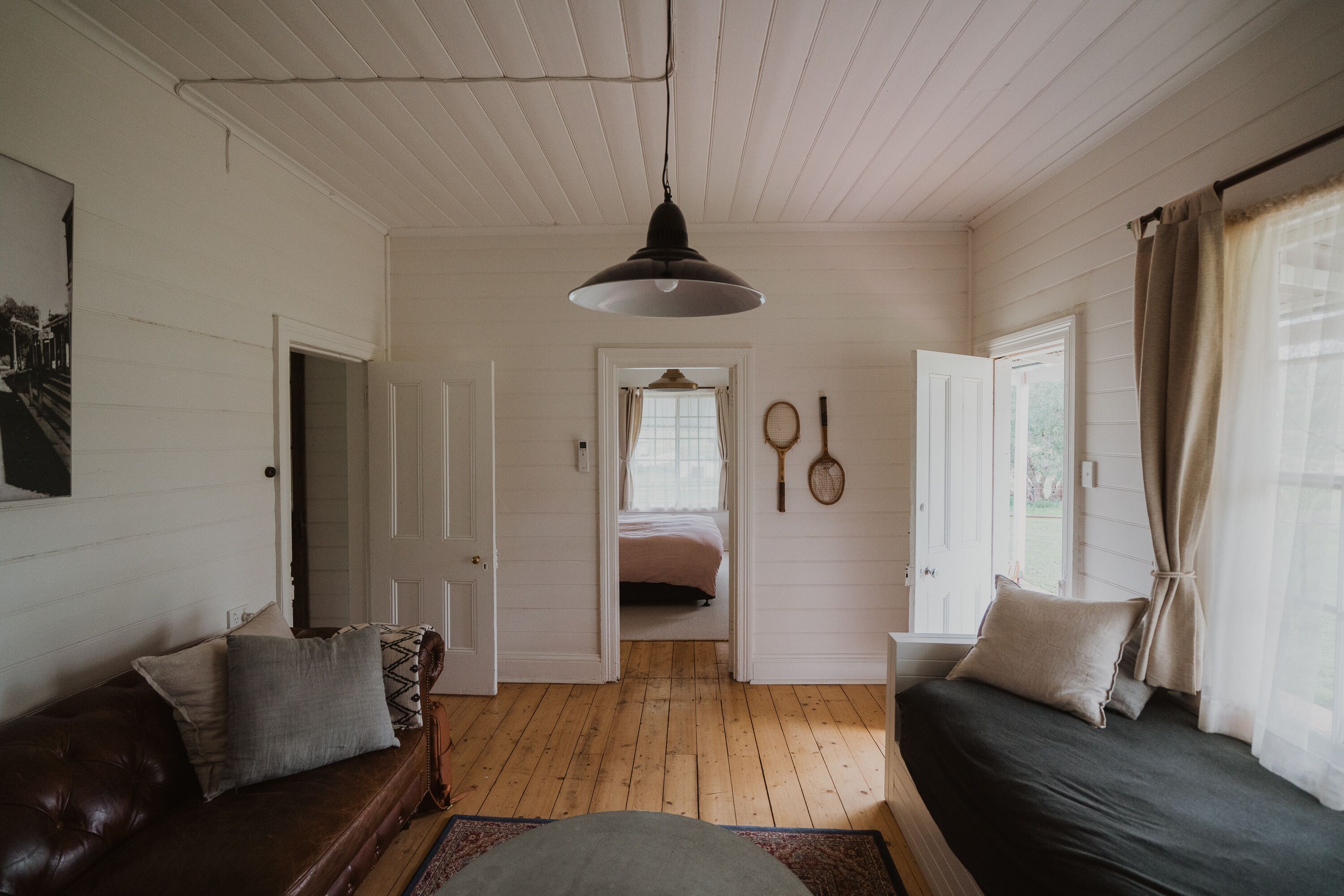 Quarto com piso de madeira, sofá de couro marrom, cama com colcha escura e luminária pendente.