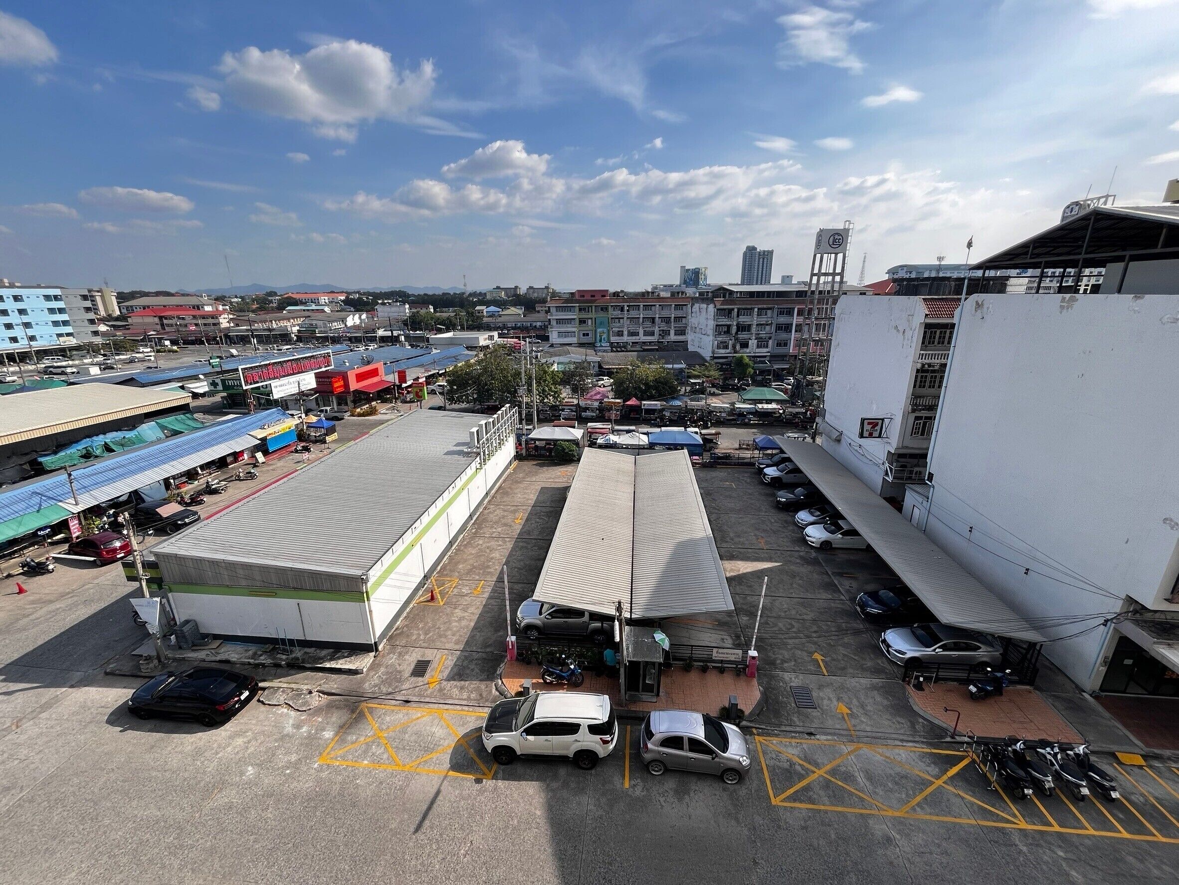 Um estacionamento com vários carros e motociclos, uma área coberta com telhado verde e edifícios ao fundo.