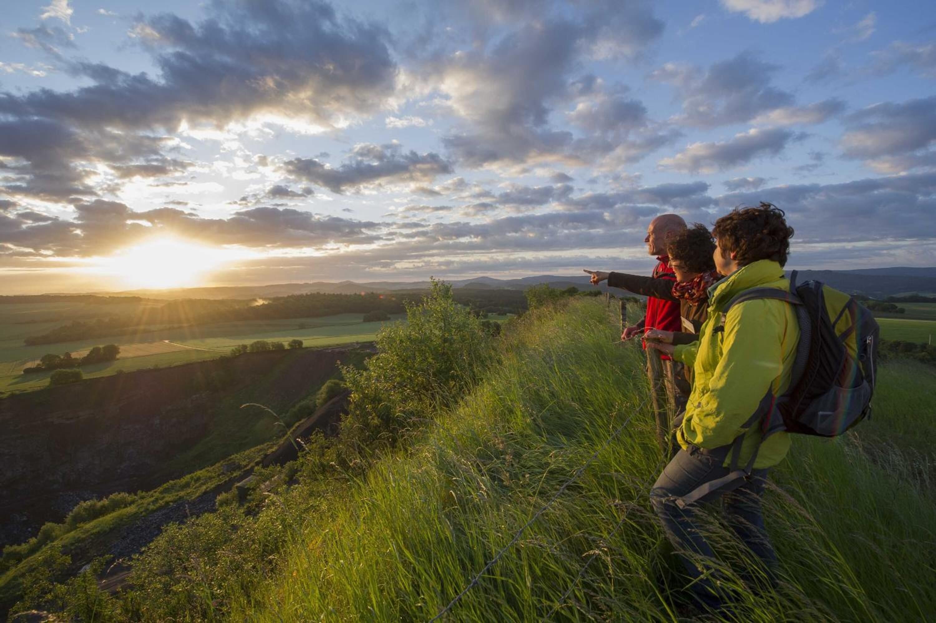 Um grupo de pessoas caminhando numa colina gramada, com um pôr do sol ao fundo.