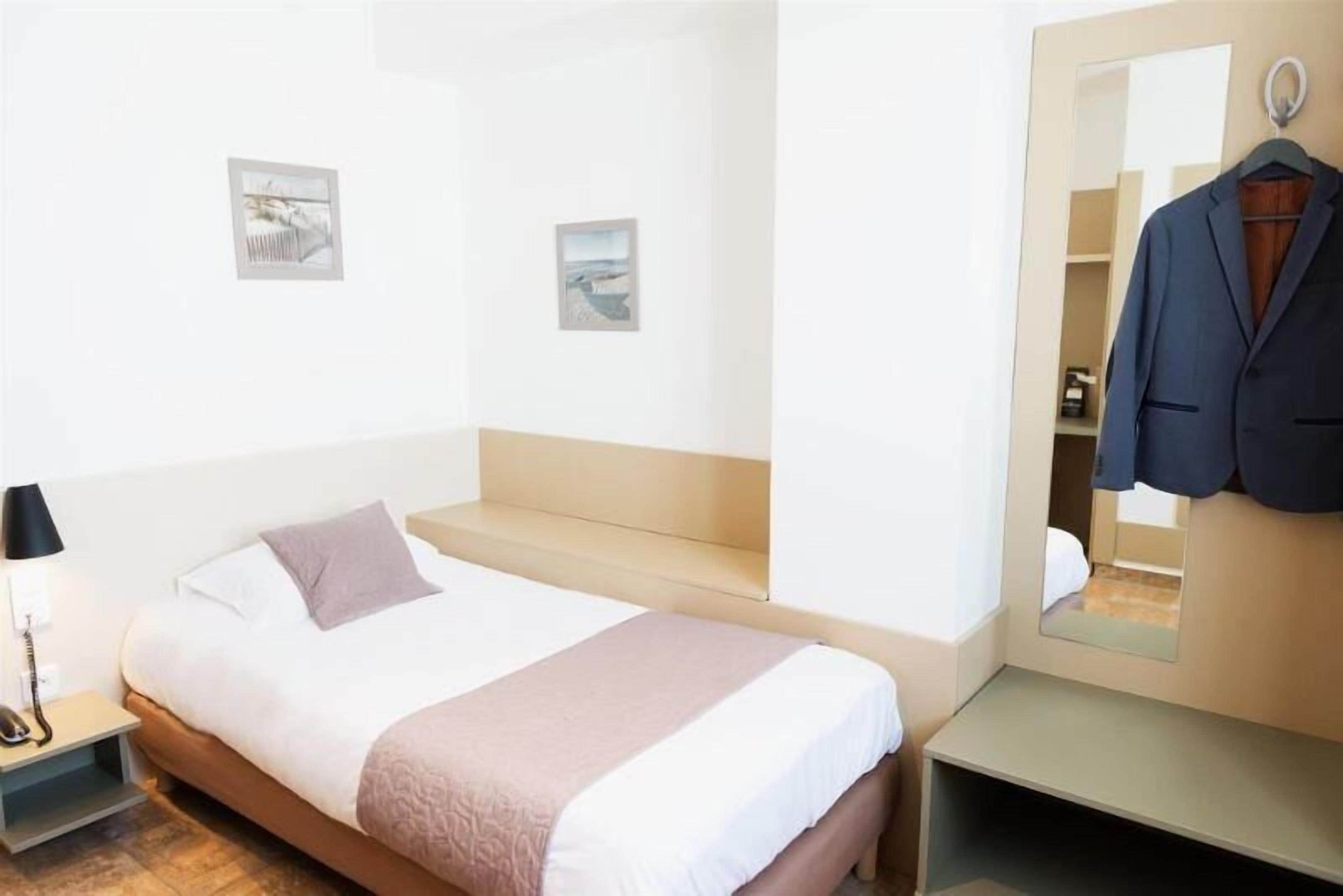 Quarto de hotel com cama, mesa de cabeceira, luminária e espelho com cabide.