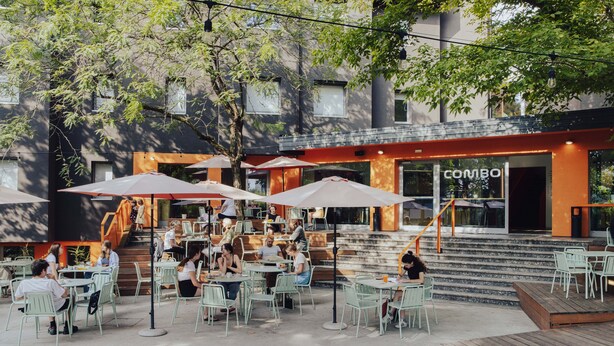 Um café ao ar livre com mesas e cadeiras, uma escadaria que leva à entrada de um edifício e uma placa com a inscrição 