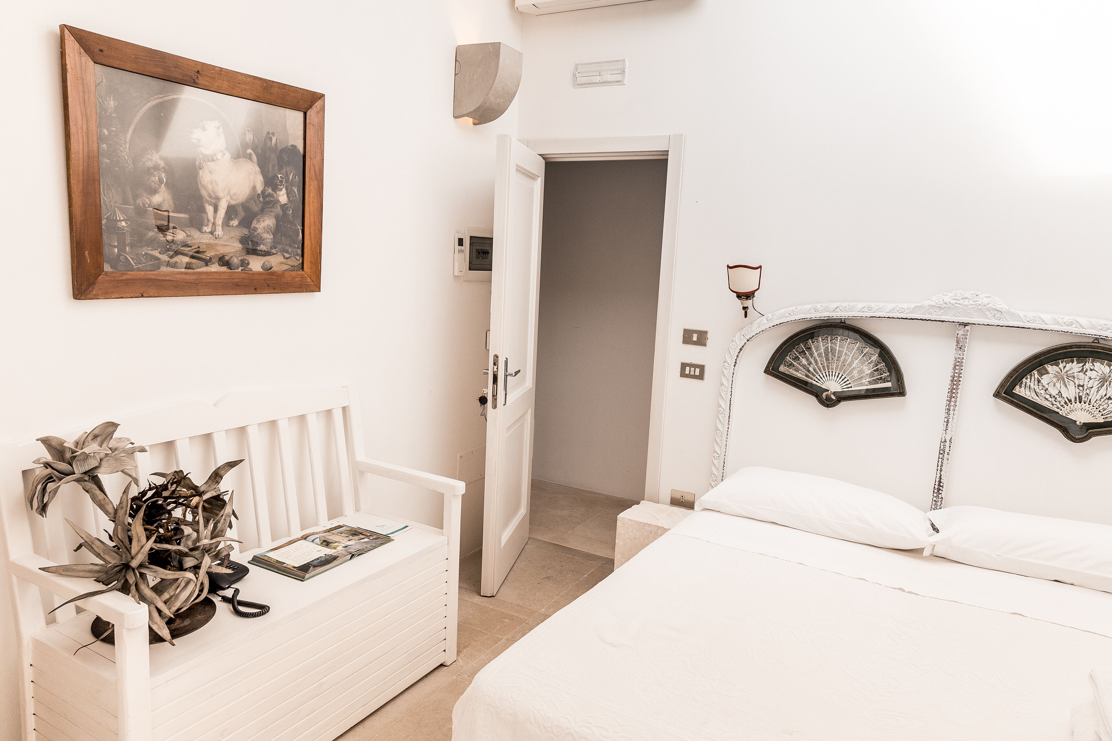 Quarto com cama branca, um banco de madeira com uma planta, um quadro na parede e uma porta que dá para outro cômodo.