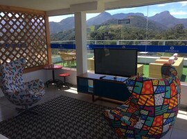 Quarto com uma grande janela que oferece vista para as montanhas, uma TV, uma cadeira colorida e uma pequena mesa com cadeiras.