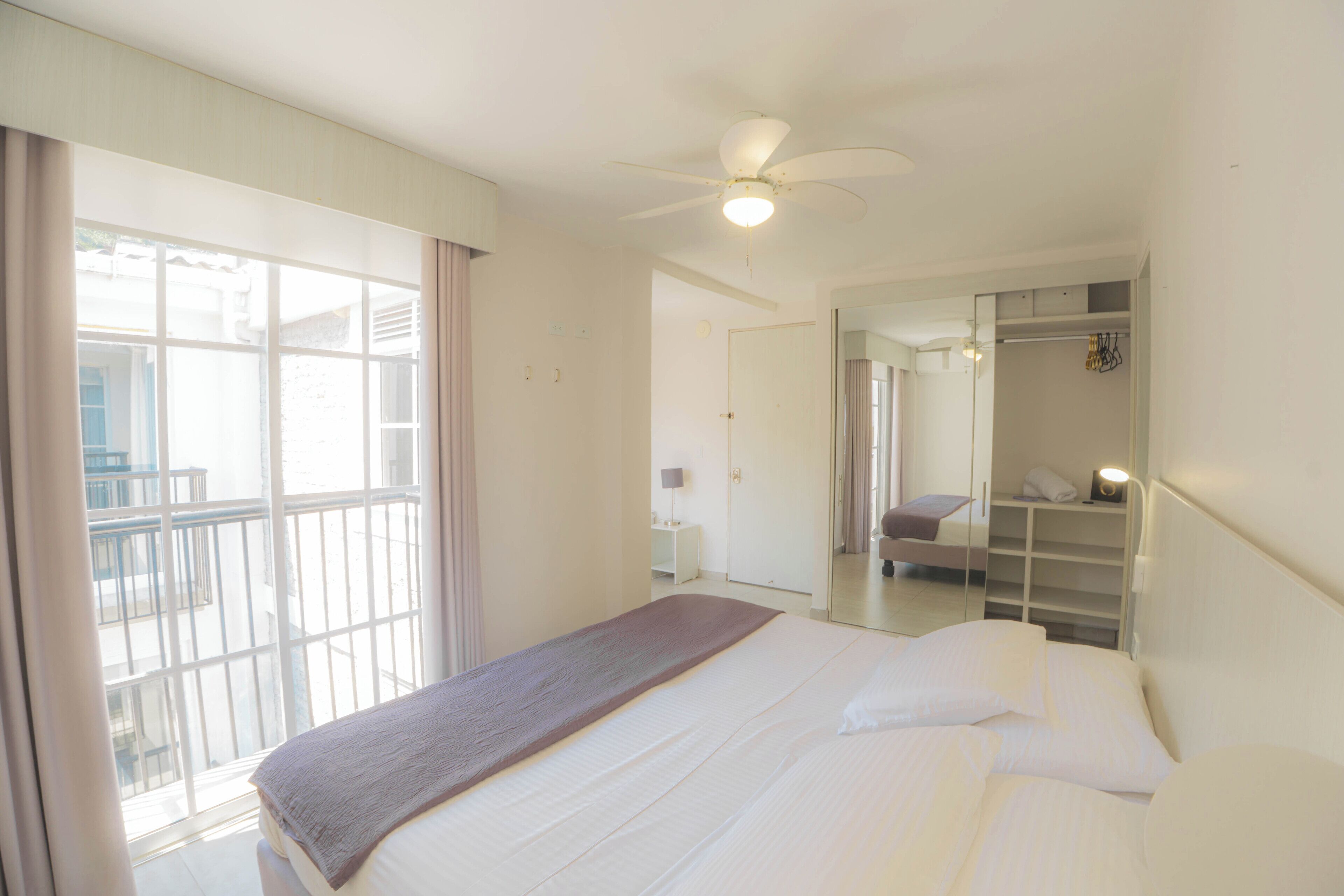 Quarto de hotel com cama, ventilador de teto, janela grande e closet espaçoso.