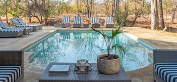 Uma área à beira da piscina com espreguiçadeiras, uma mesa com um livro e uma planta, e vista para árvores ao fundo.