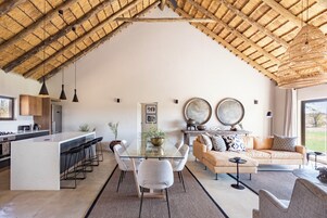 Cozinha moderna com ilha central, banquetas de bar e mesa de jantar com cadeiras. Possui um sofá e uma mesa de centro na área de estar, com uma janela ampla que oferece vista para o exterior.