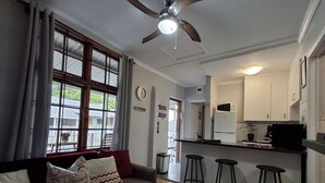 Uma sala de estar com sofá marrom, ventilador de teto e área de cozinha com geladeira e banquetas de bar.