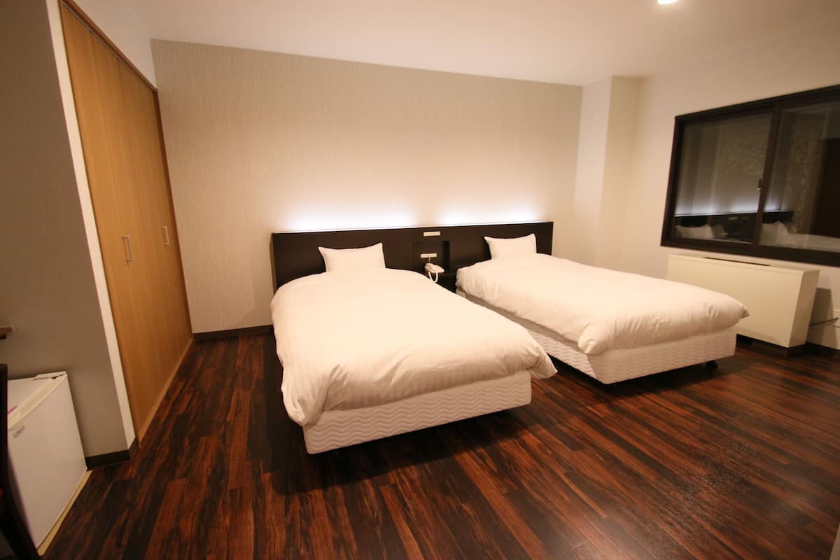 Quarto de hotel com duas camas de solteiro, piso de madeira, uma janela grande e um guarda-roupa.