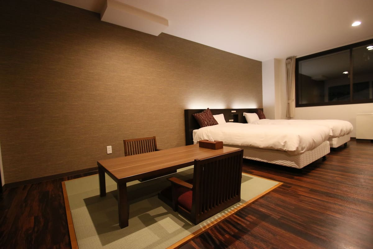 Quarto de hotel com piso de madeira, cama com roupa de cama branca, uma mesa de madeira e uma cadeira.