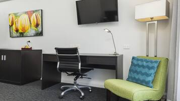 Quarto de hotel com escrivaninha, cadeira, televisão fixada na parede, luminária e quadro de flores.