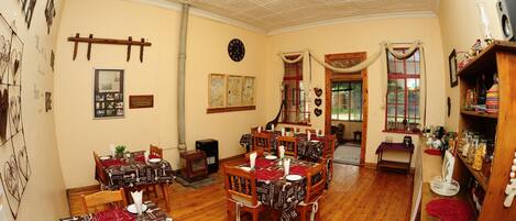 Uma área de refeições com mesas e cadeiras de madeira, um relógio na parede e uma estante com diversos objetos.