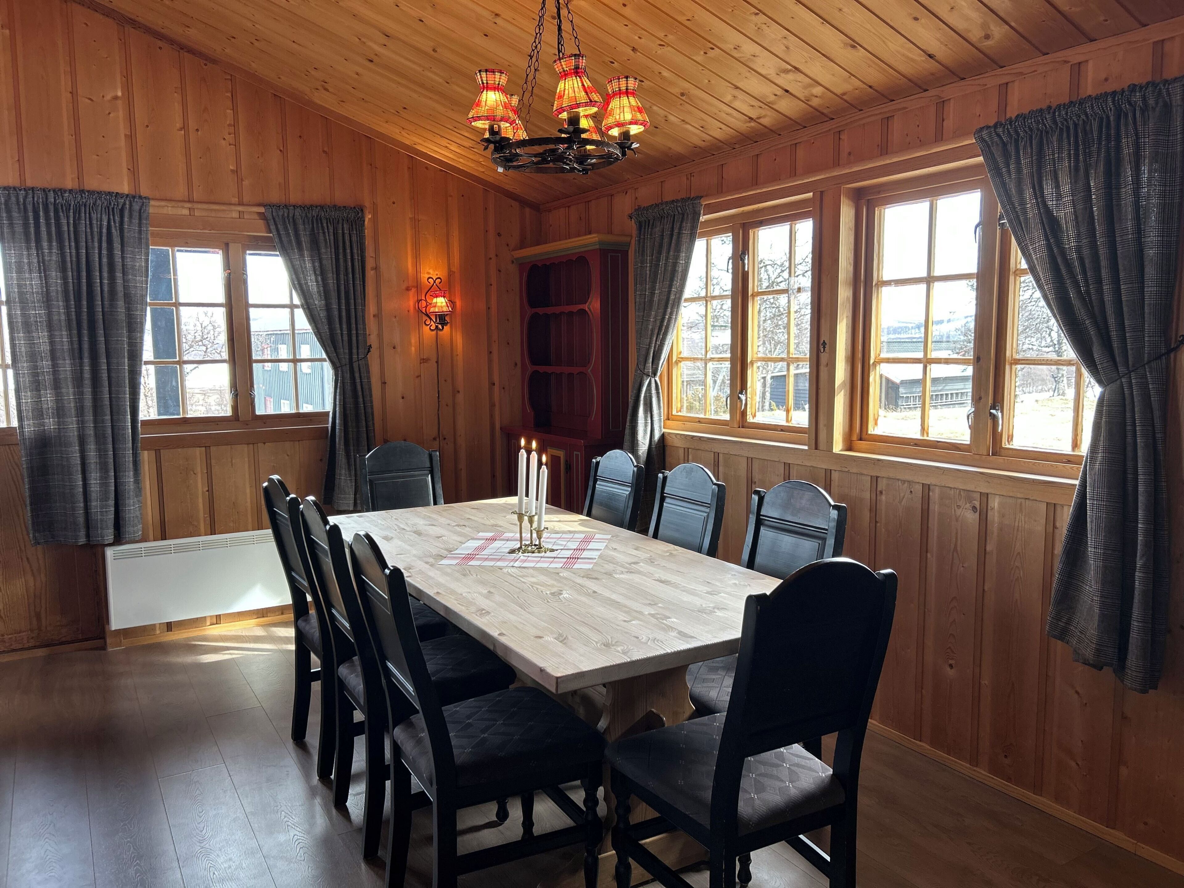Um quarto com painéis de madeira, uma mesa de jantar, cadeiras, um lustre e grandes janelas com cortinas.