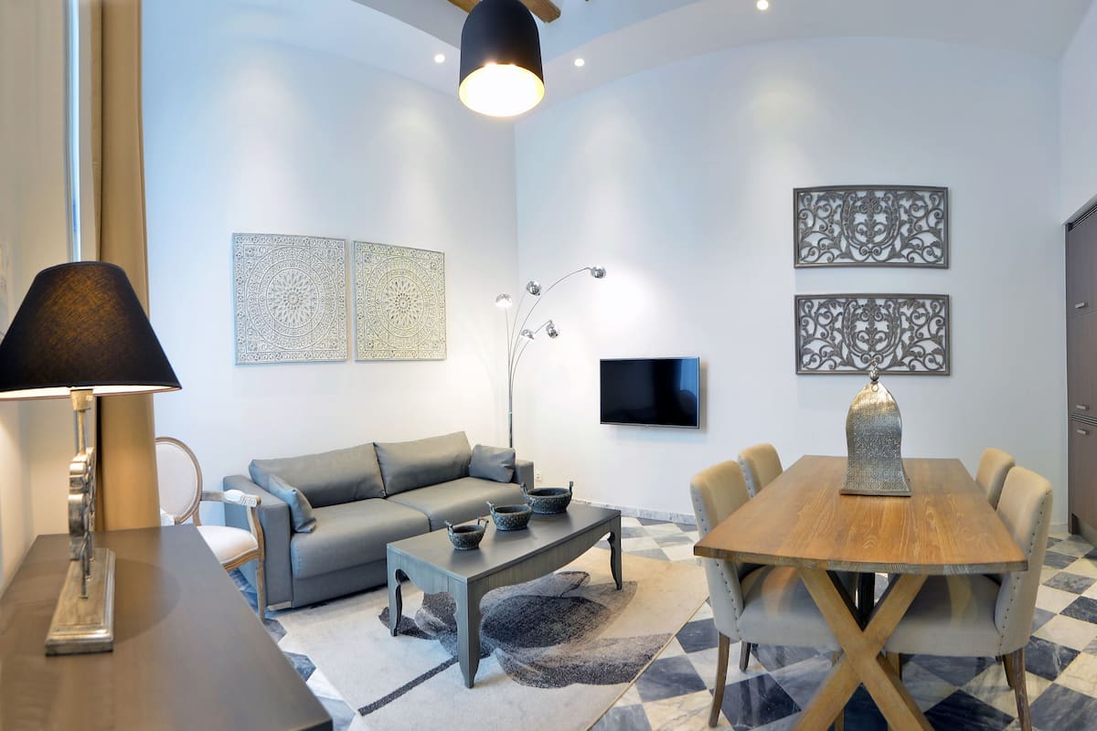 Uma sala de estar moderna com sofá, mesa de centro e televisão. Inclui uma mesa de jantar com cadeiras e uma cozinha com armários e pia.