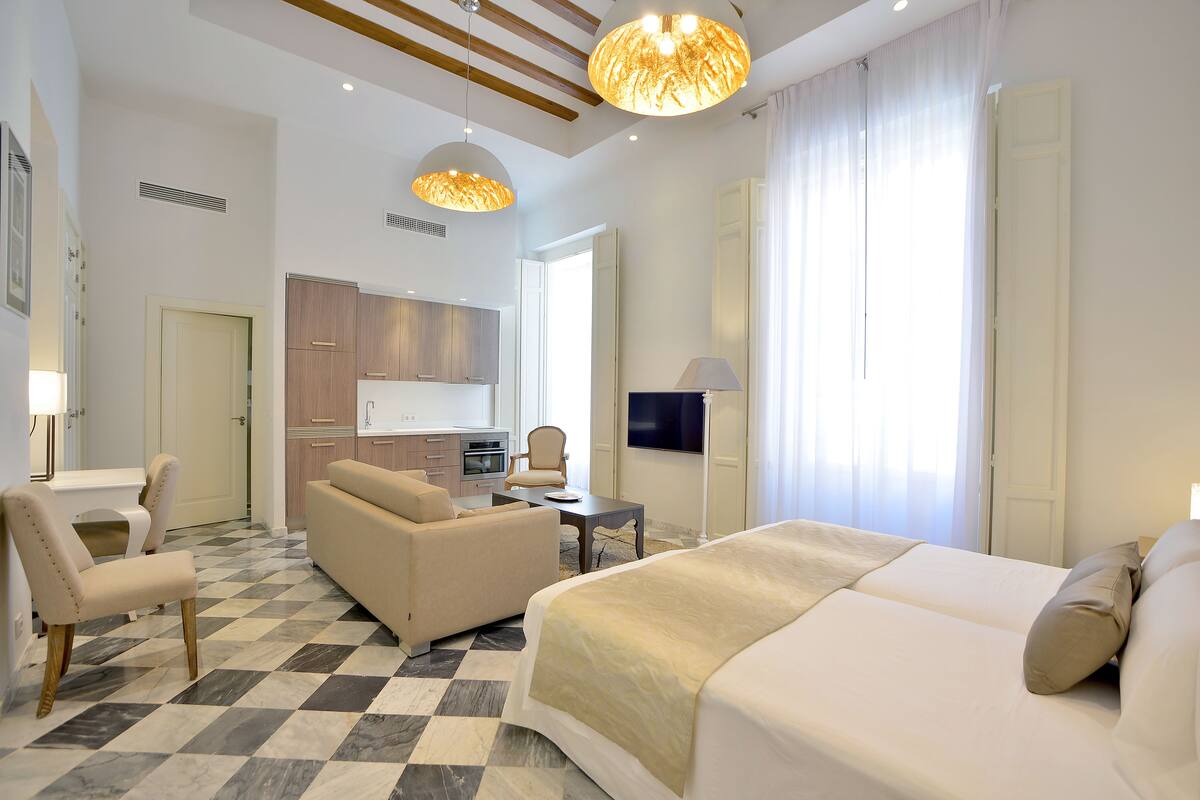 Quarto de hotel com cama, sofá, poltrona, escrivaninha e cozinha compacta.