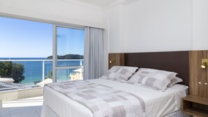 Quarto com uma cama grande, cabeceira de madeira e vista para o mar.