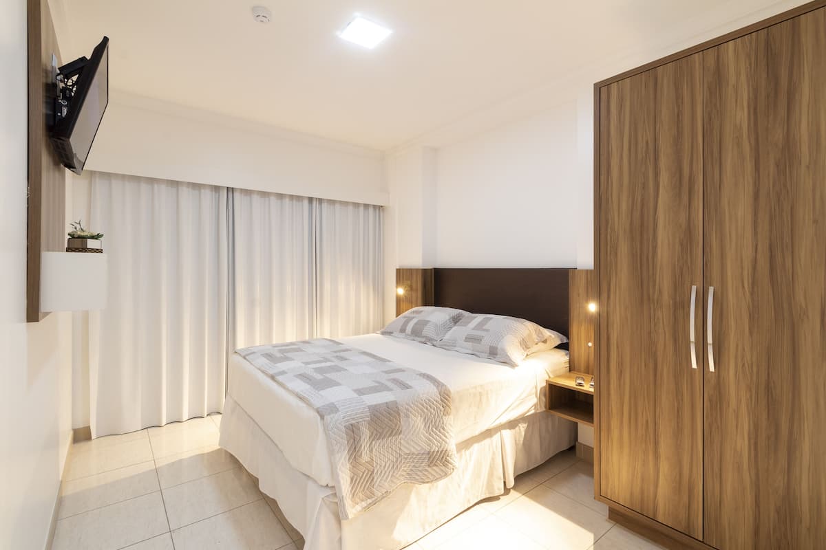 Quarto moderno com uma cama, um guarda-roupa de madeira, uma mesa de cabeceira e uma TV fixada na parede.