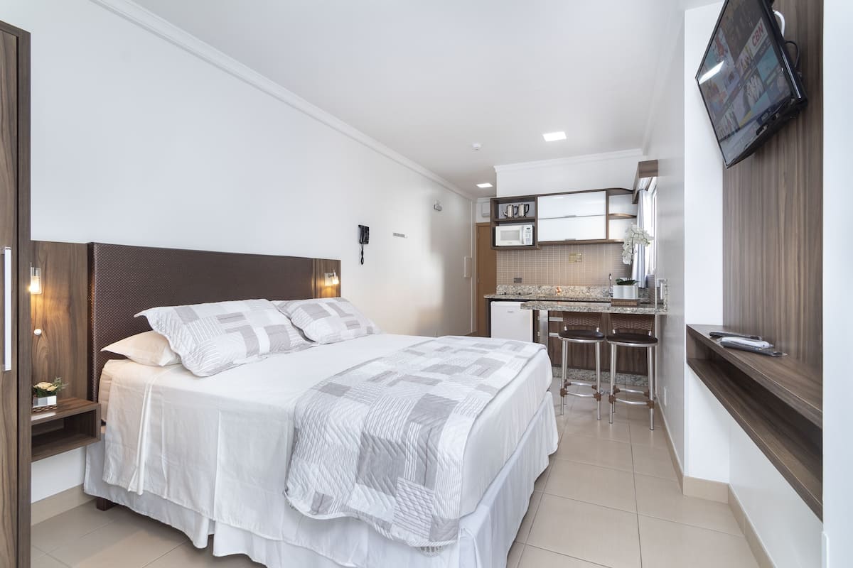 Quarto de hotel moderno com uma cama bem arrumada, uma pequena cozinha e uma TV fixada na parede.
