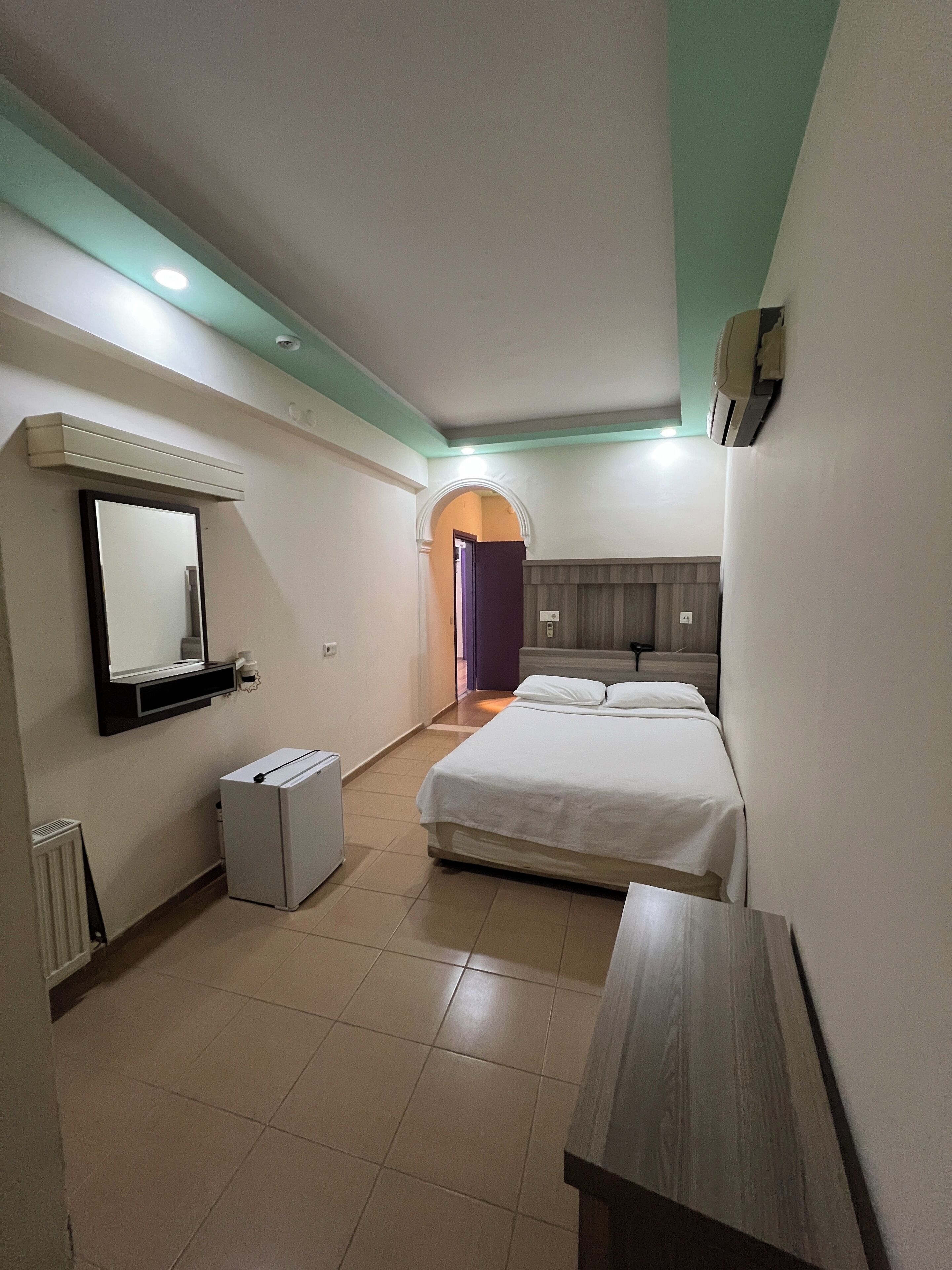 Quarto de hotel com cama, uma mesa pequena, espelho, radiador e porta que dá para outro cômodo.