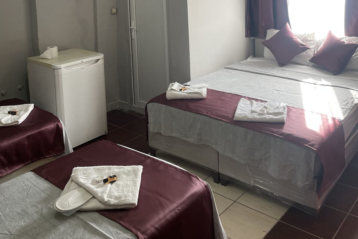 Quarto de hotel com duas camas, um ventilador de teto, uma janela com cortinas e um eletrodoméstico branco pequeno.
