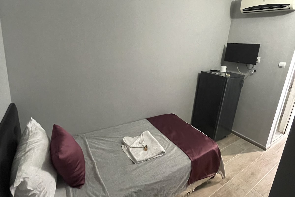 Quarto pequeno com uma cama de solteiro, parede cinzenta, piso de madeira, televisão fixada na parede e ar condicionado sobre a cama.