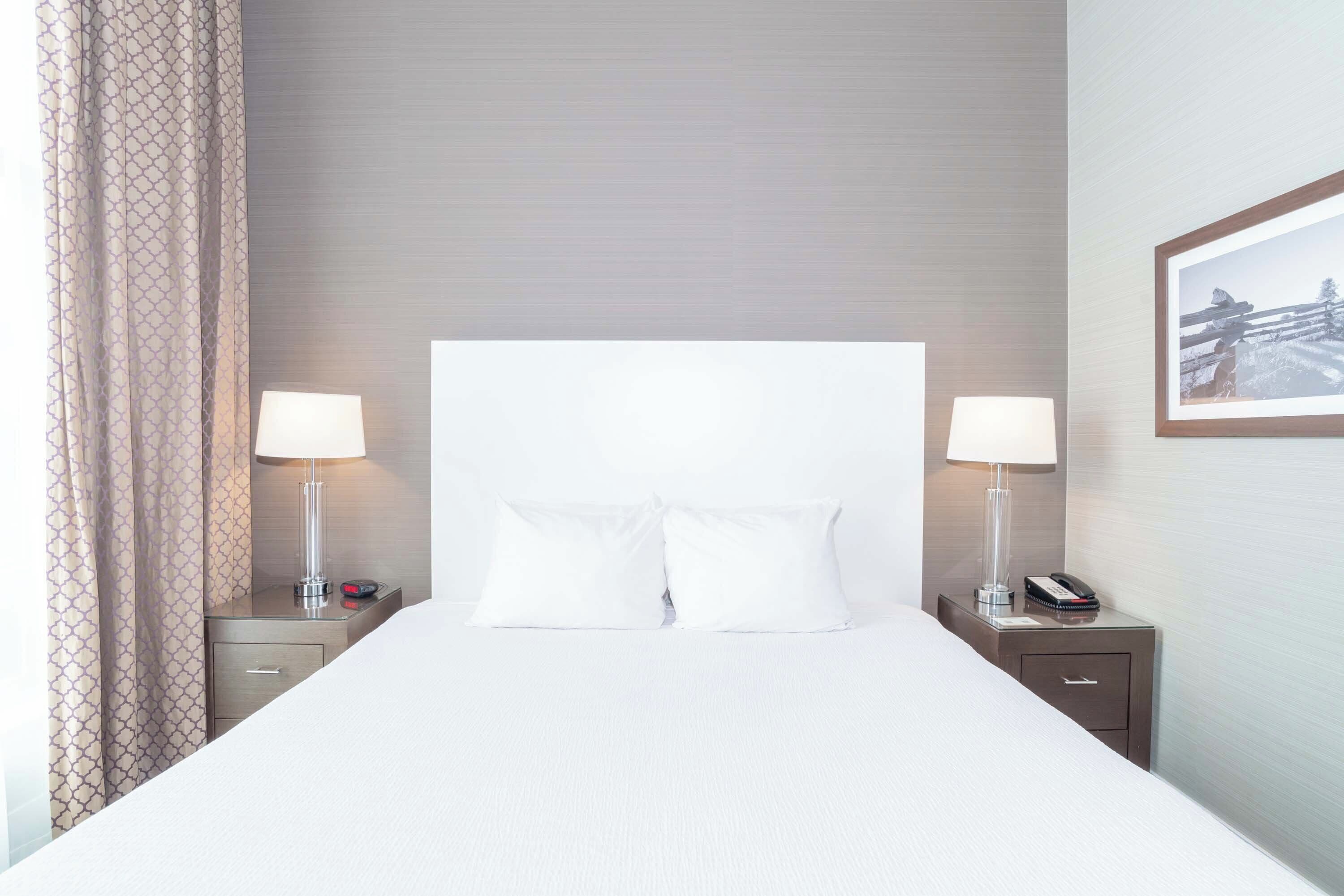 Quarto de hotel com uma cama grande, duas mesas de cabeceira com abajures, um quadro na parede e uma cortina estampada.