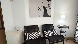 Quarto com duas poltronas de couro preto com almofadas listradas, um armário branco pequeno, uma mesa redonda com abajur e um quadro abstrato grande na parede.