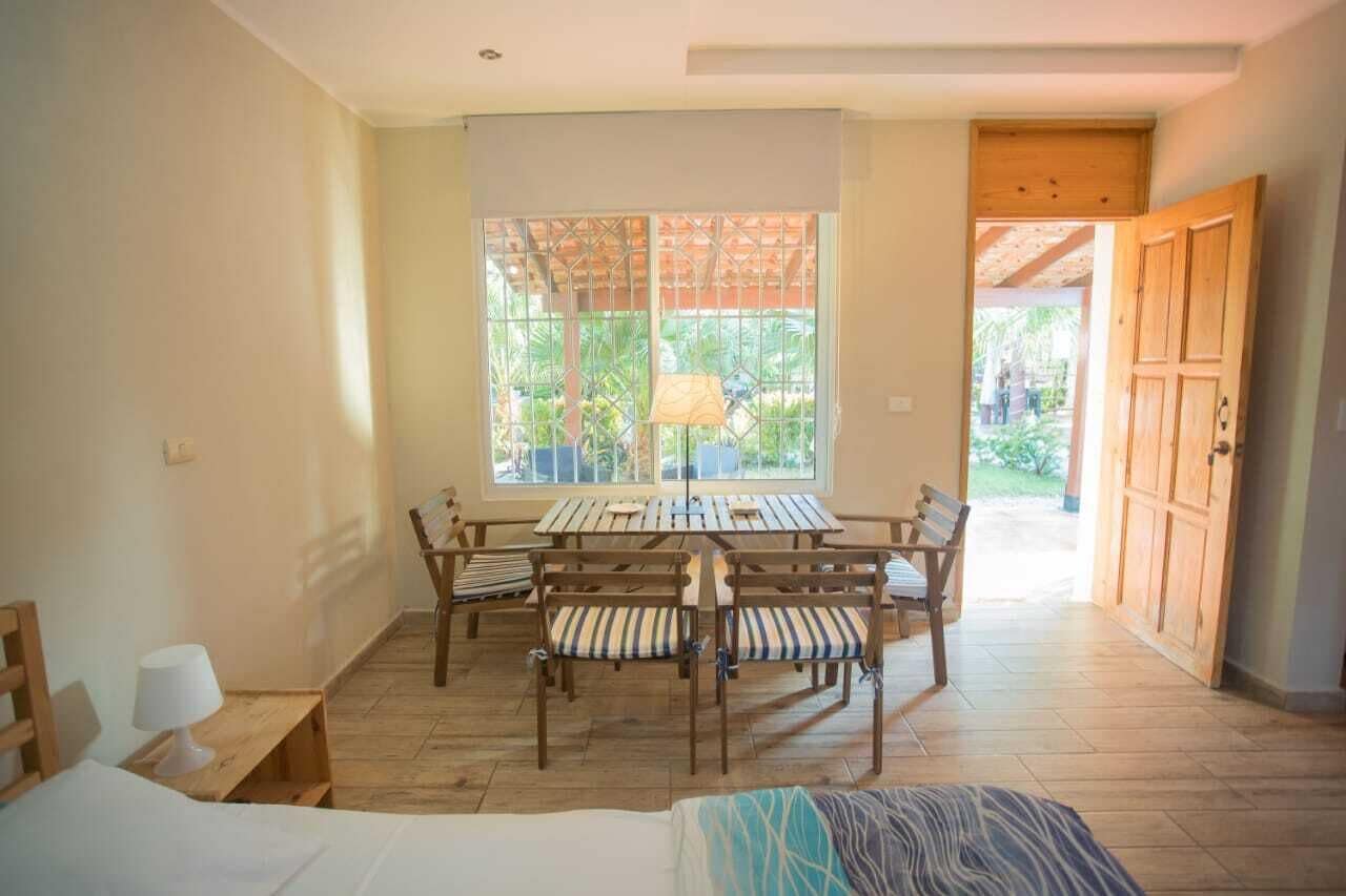 Quarto com cama, mesa e cadeiras, janela com vista para o verde e porta que dá para o exterior.