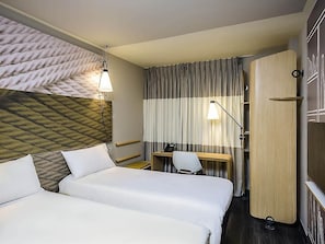 Quarto de hotel com duas camas, uma escrivaninha, uma cadeira, um guarda-roupa e cabeceira com padrão decorativo.