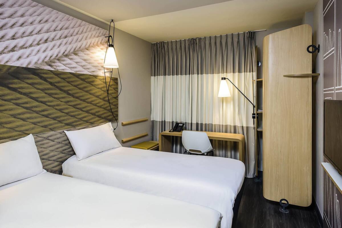 Quarto de hotel com duas camas, uma escrivaninha, uma cadeira, um guarda-roupa e cabeceira com padrão decorativo.