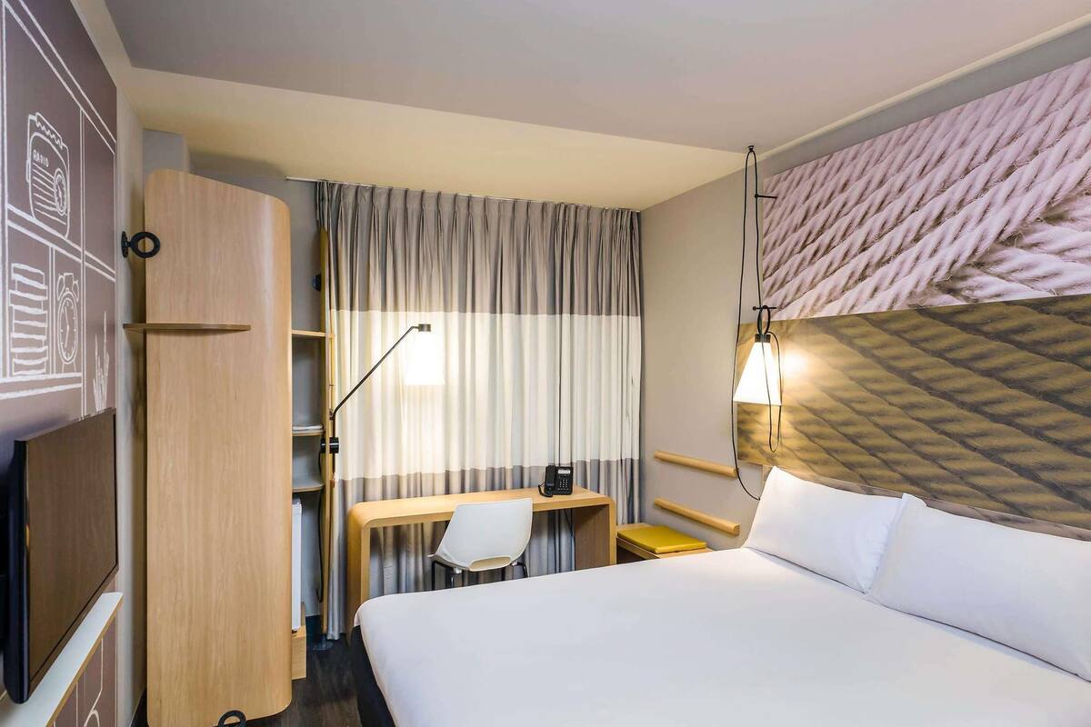 Quarto de hotel com cama, escrivaninha, cadeira, guarda-roupa e luminária fixada na parede.