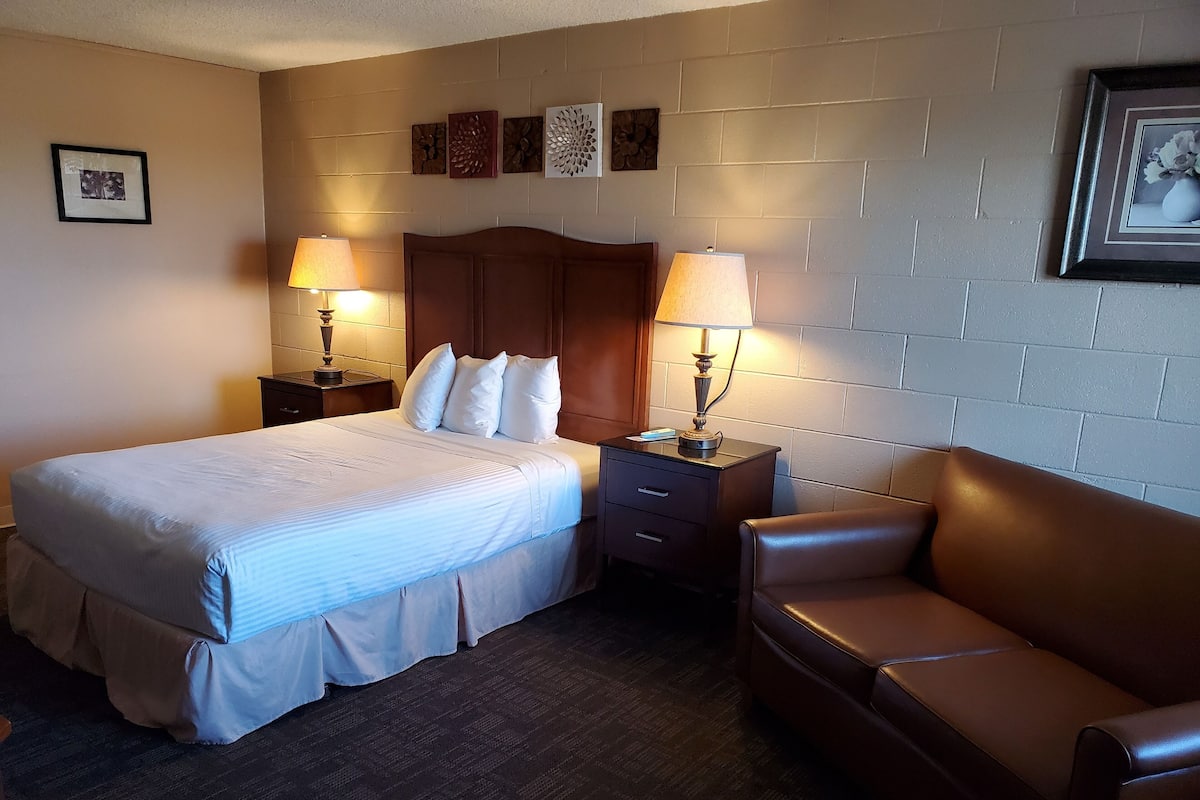 Quarto de hotel com uma cama grande, duas mesas de cabeceira com luminárias, um sofá de couro marrom e uma parede com padrão de tijolos com obras de arte emolduradas.